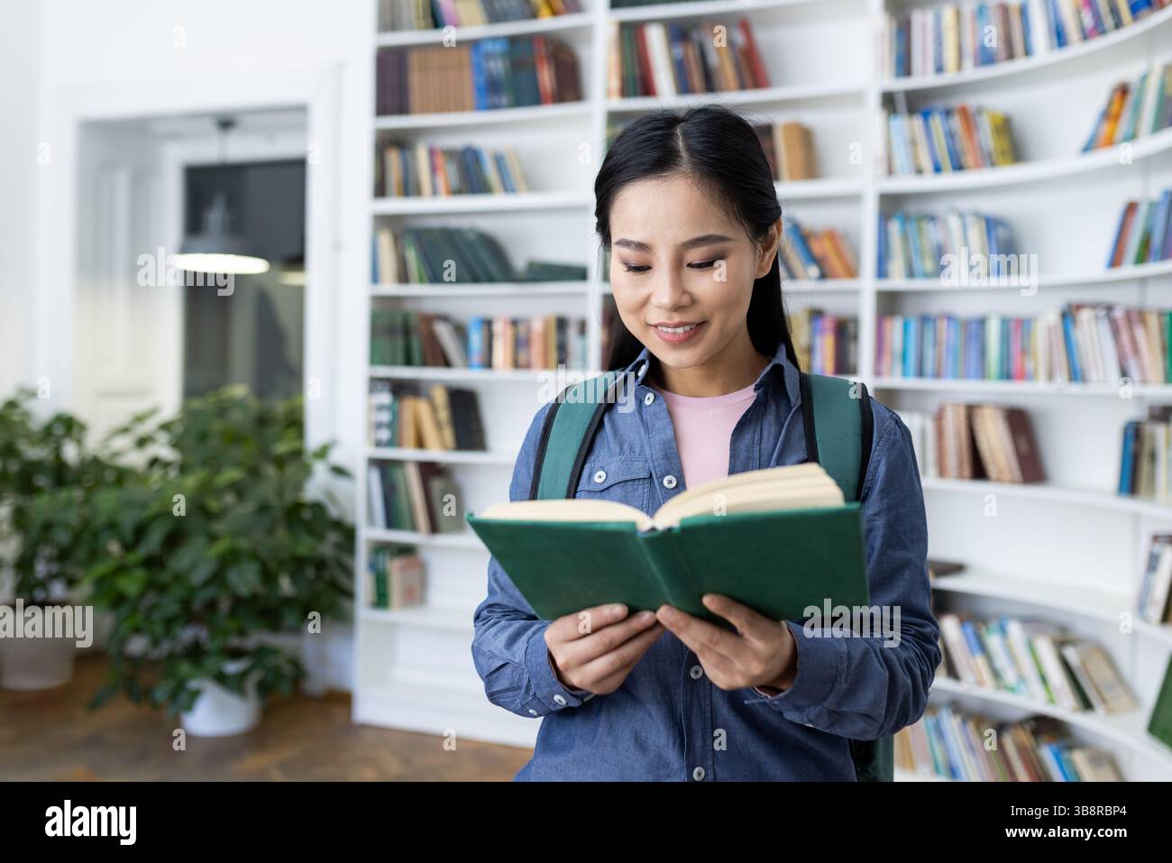 A young Asian woman smiles as she reads a book in a library, surrounded ...