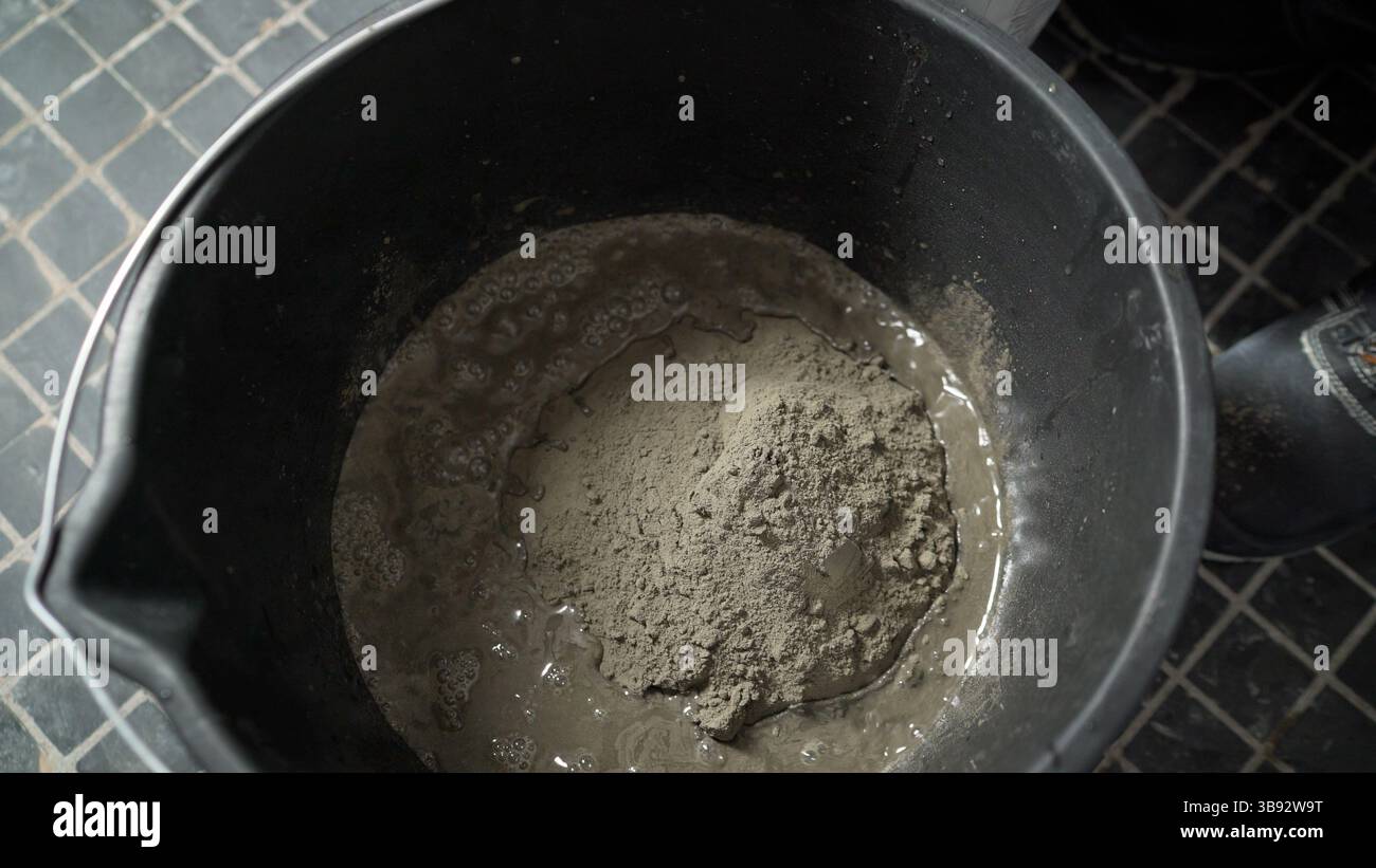 Cement powder is being mixed with water in a black bucket, creating a ...