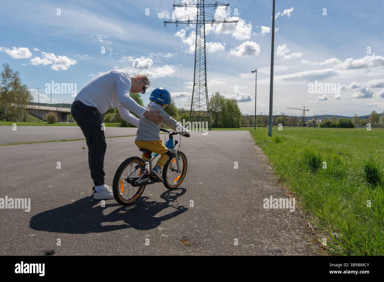 Parent supports child learning to ride a bicycle on an open road under ...