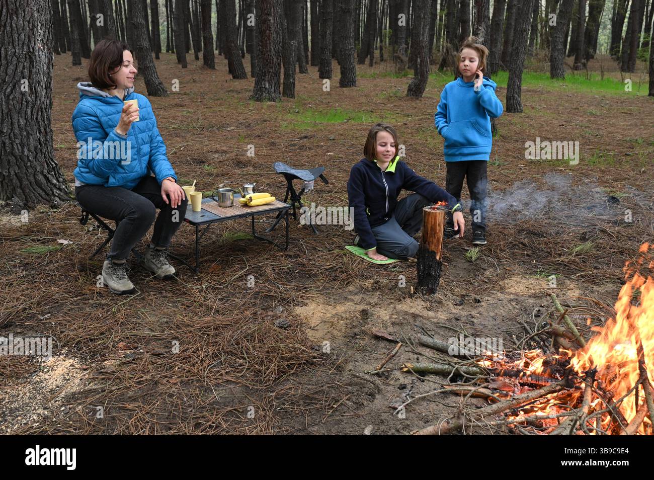 Mother and two children sit by a campfire in the forest, eating and ...