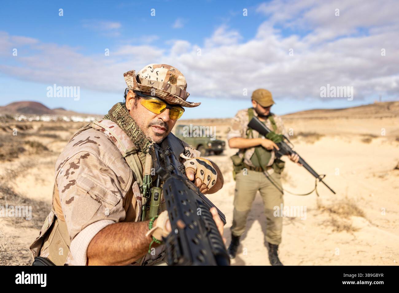 Two soldiers in camouflage gear conduct training in a desert landscape, aiming rifles with ...