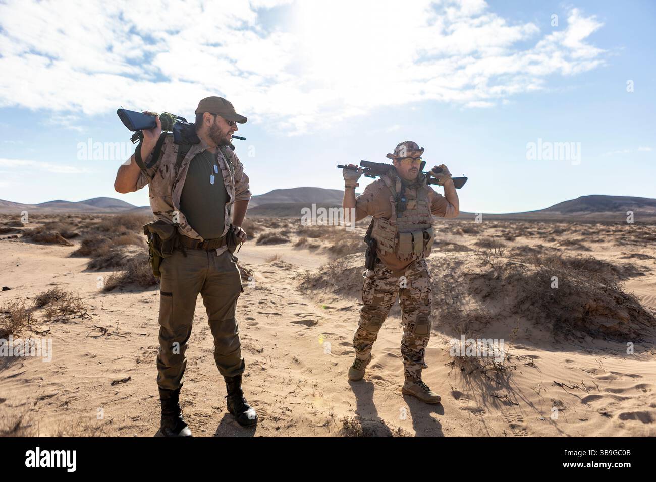 Two soldiers in tactical gear patrol a sunny desert landscape. They ...