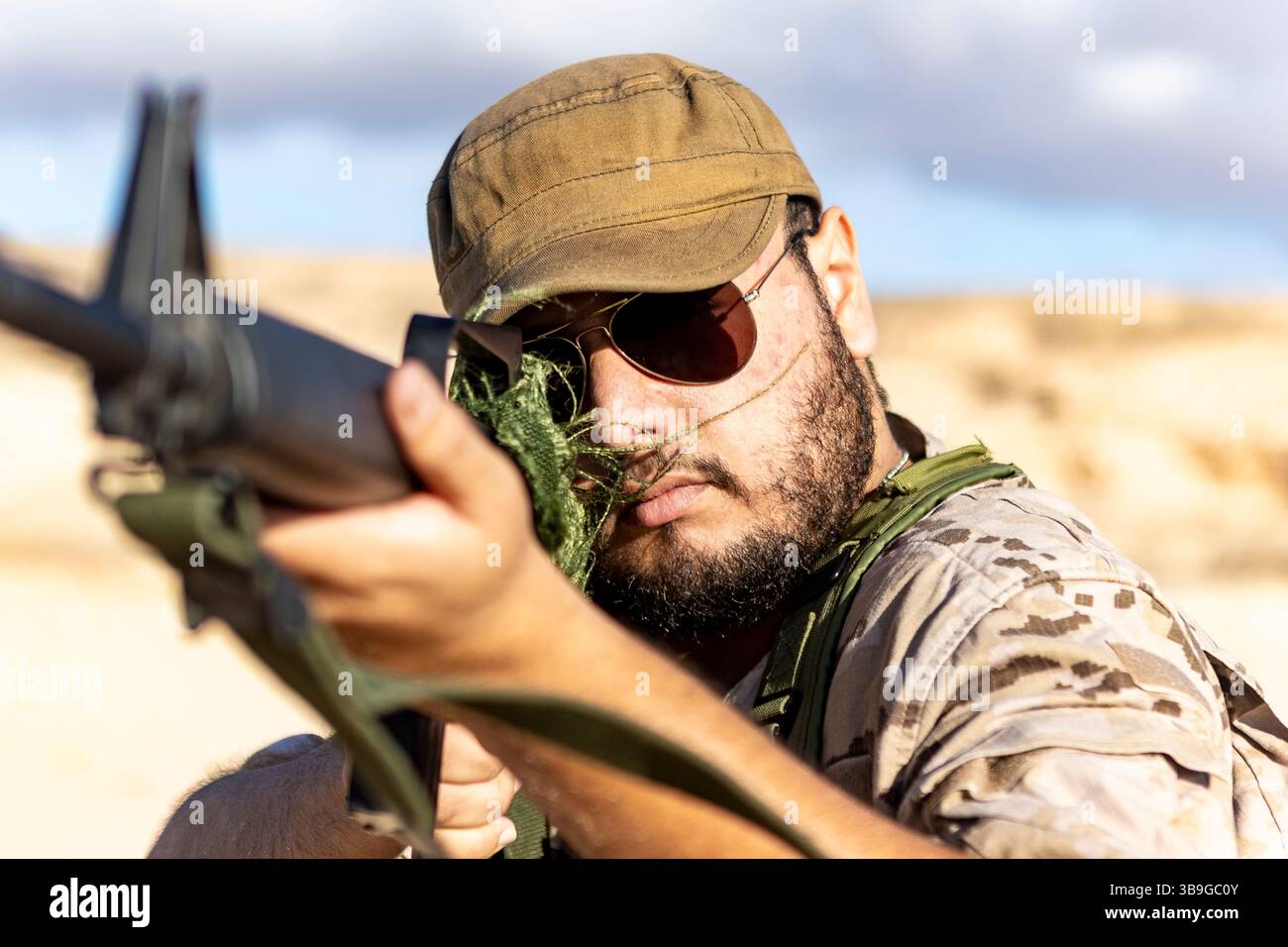 A soldier in camouflage uniform aiming a rifle in a desert landscape ...