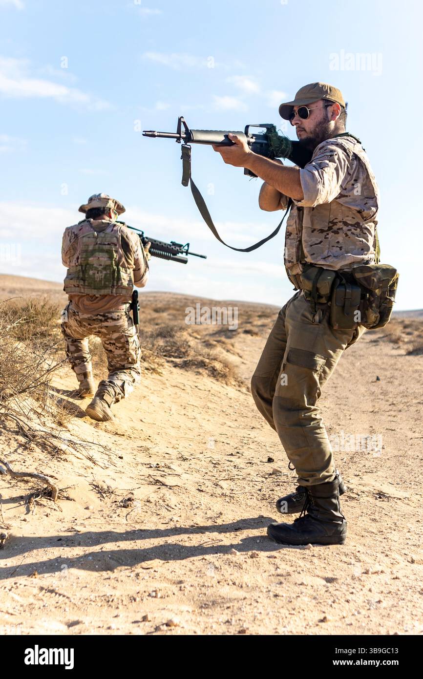 Two soldiers in tactical gear advance through a desert. They are ...