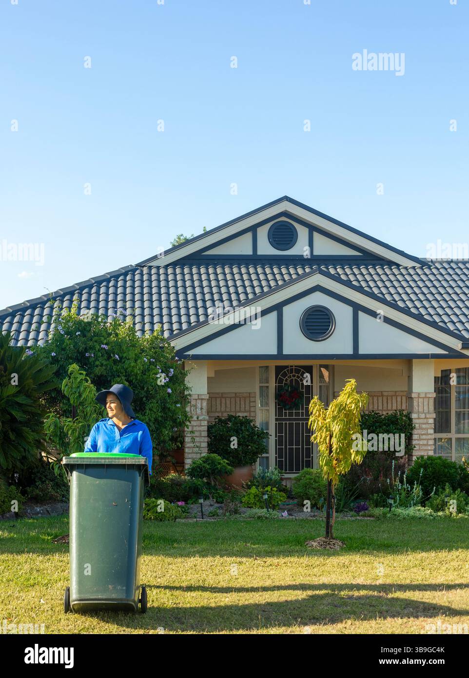A woman pushes a garbage bin outside a typical Australian suburban ...