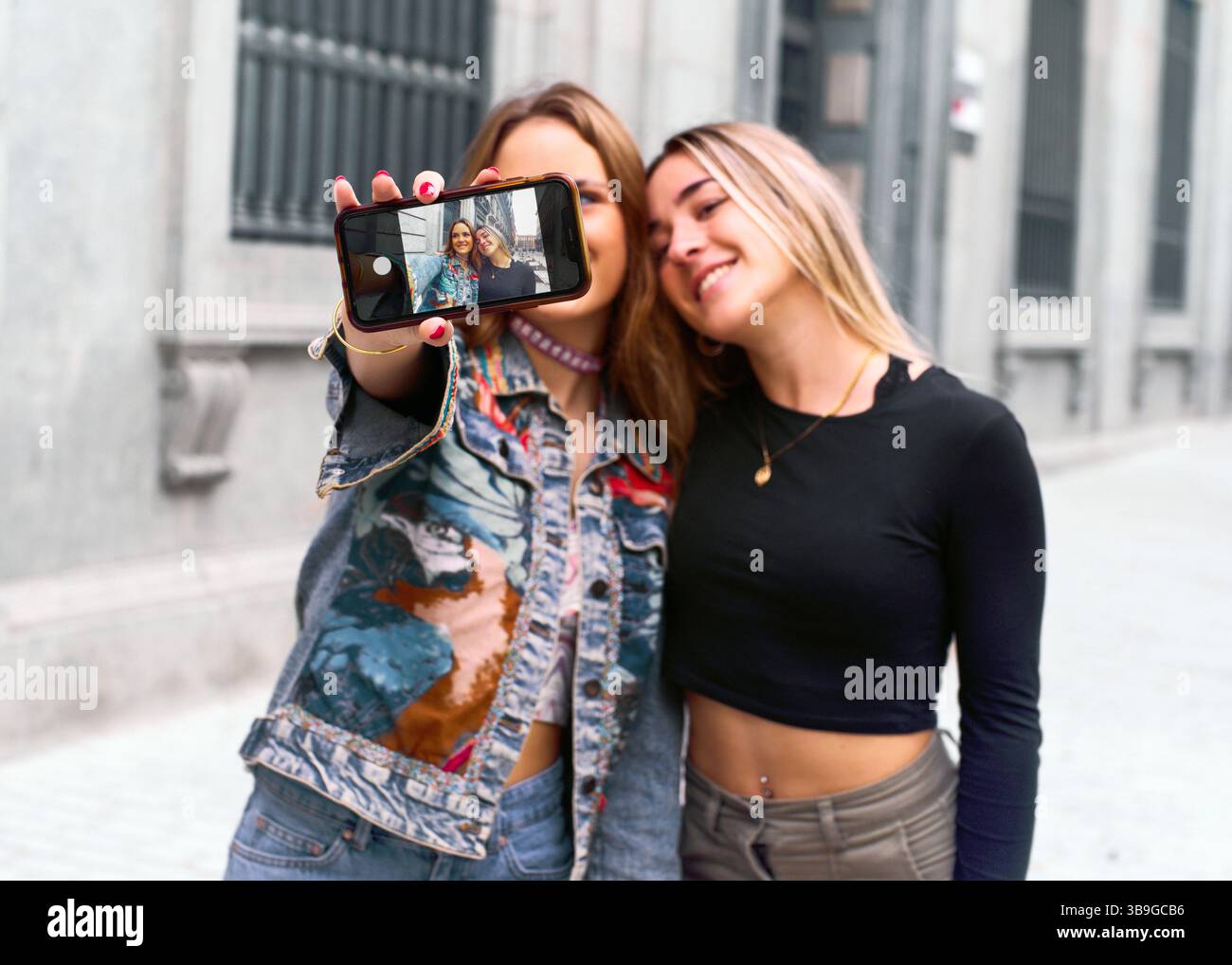 Two young friends pose for a selfie outdoors in an urban setting. One ...