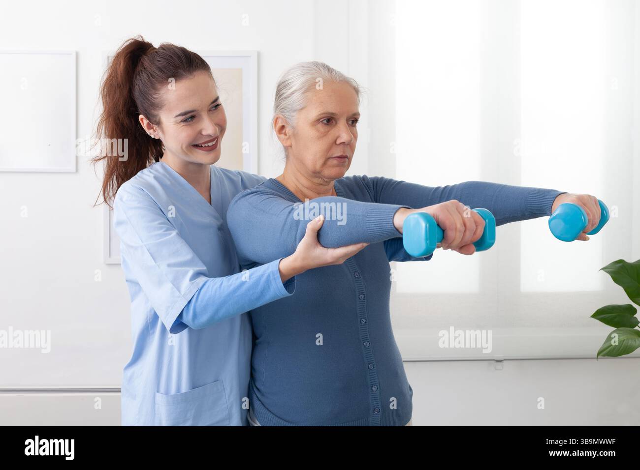 Smiling young nurse assisting an elderly woman with light dumbbell ...