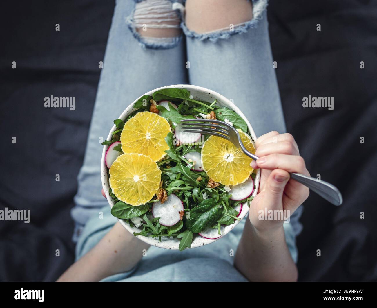 Woman in jeans at bed, holding vegan salad bowl with oranges, spinach ...