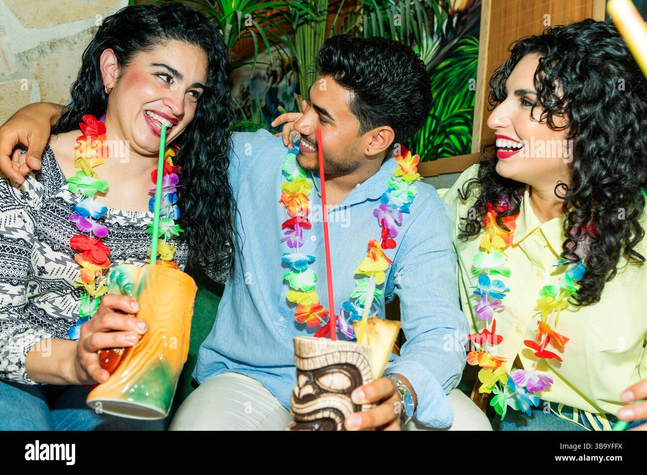 Three cheerful friends wearing hawaiian leis are having fun, drinking ...