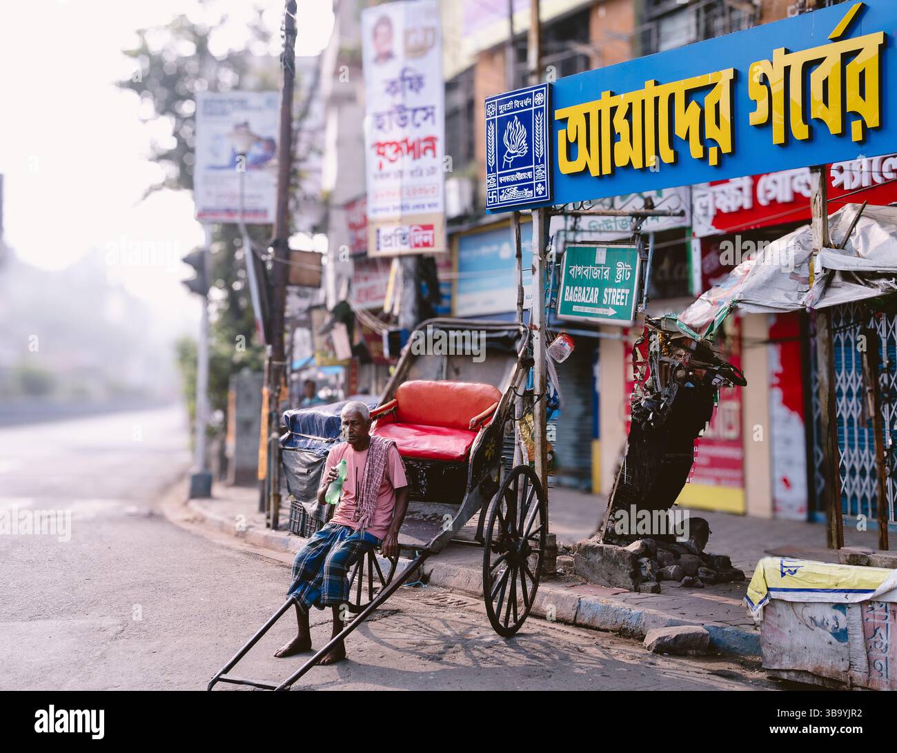 An aged hand-pulled rickshaw puller rests on his rickshaw along a busy ...