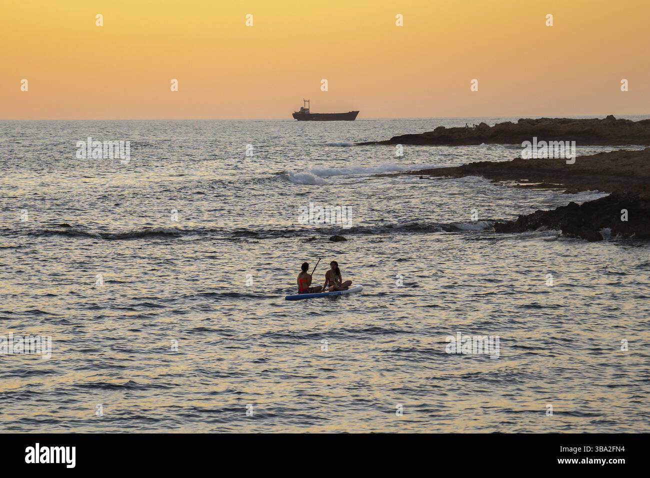 Two girls sit on sup board in the quiet mediterranean sea at sunset in ...