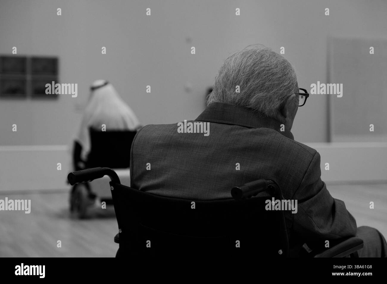 Two elderly figures in wheelchairs sit silently in the exhibit space ...