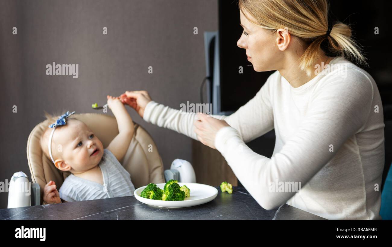 Mother trying to feed baby with spoon healthy broccoli at home, toddler ...