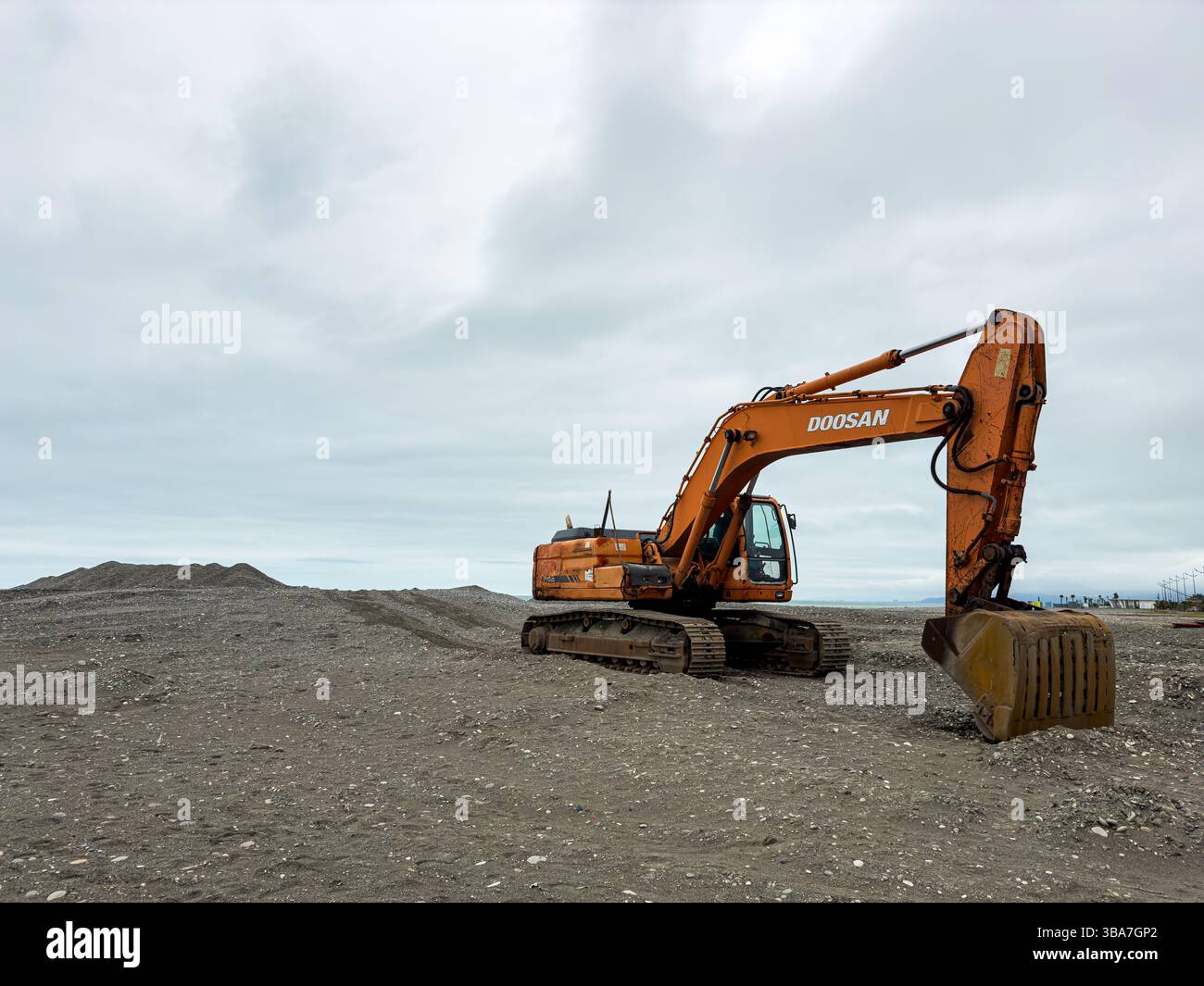 Excavator on deserted shoreline construction site under overcast sky. Wide shot, no people ...