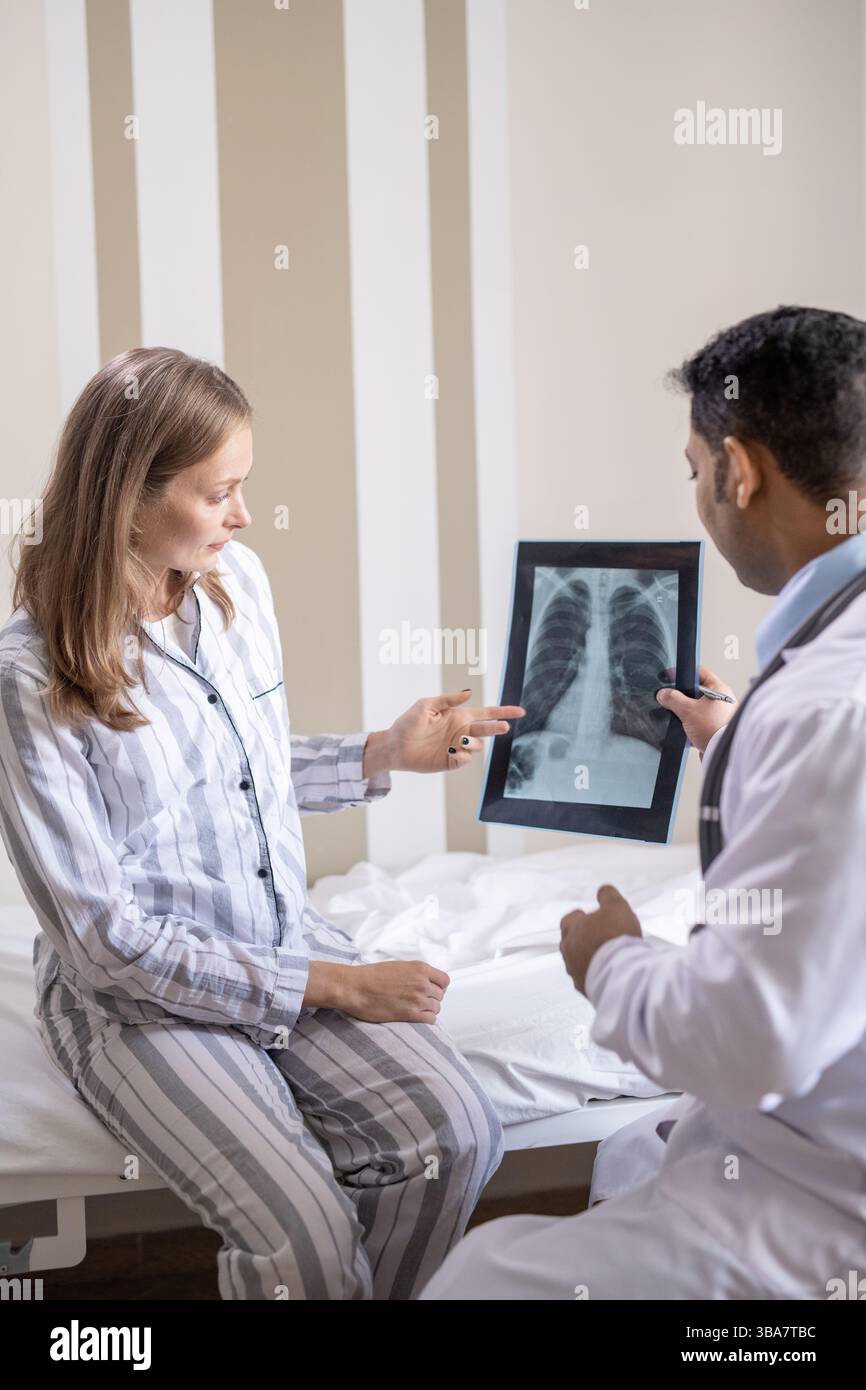 Young female in pajamas sitting on bed in front of her doctor during ...