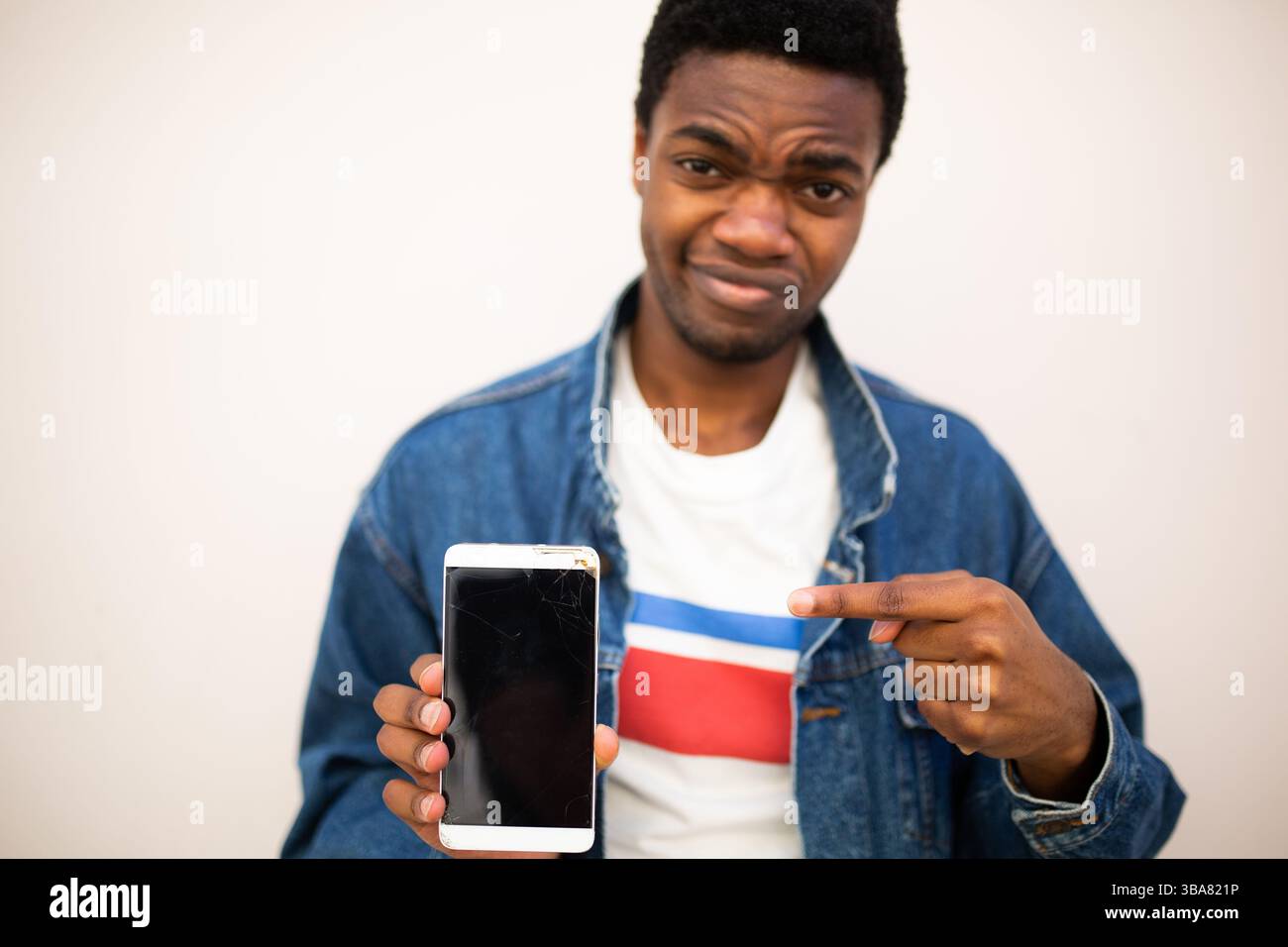 Black male pointing finger on cellphone standing against white wall ...