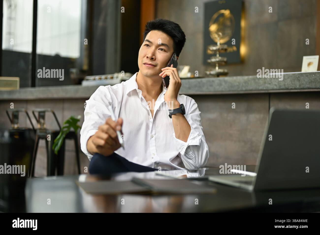 Handsome millennial Asian businessman calling someone, talking on the phone while sitting in a ...