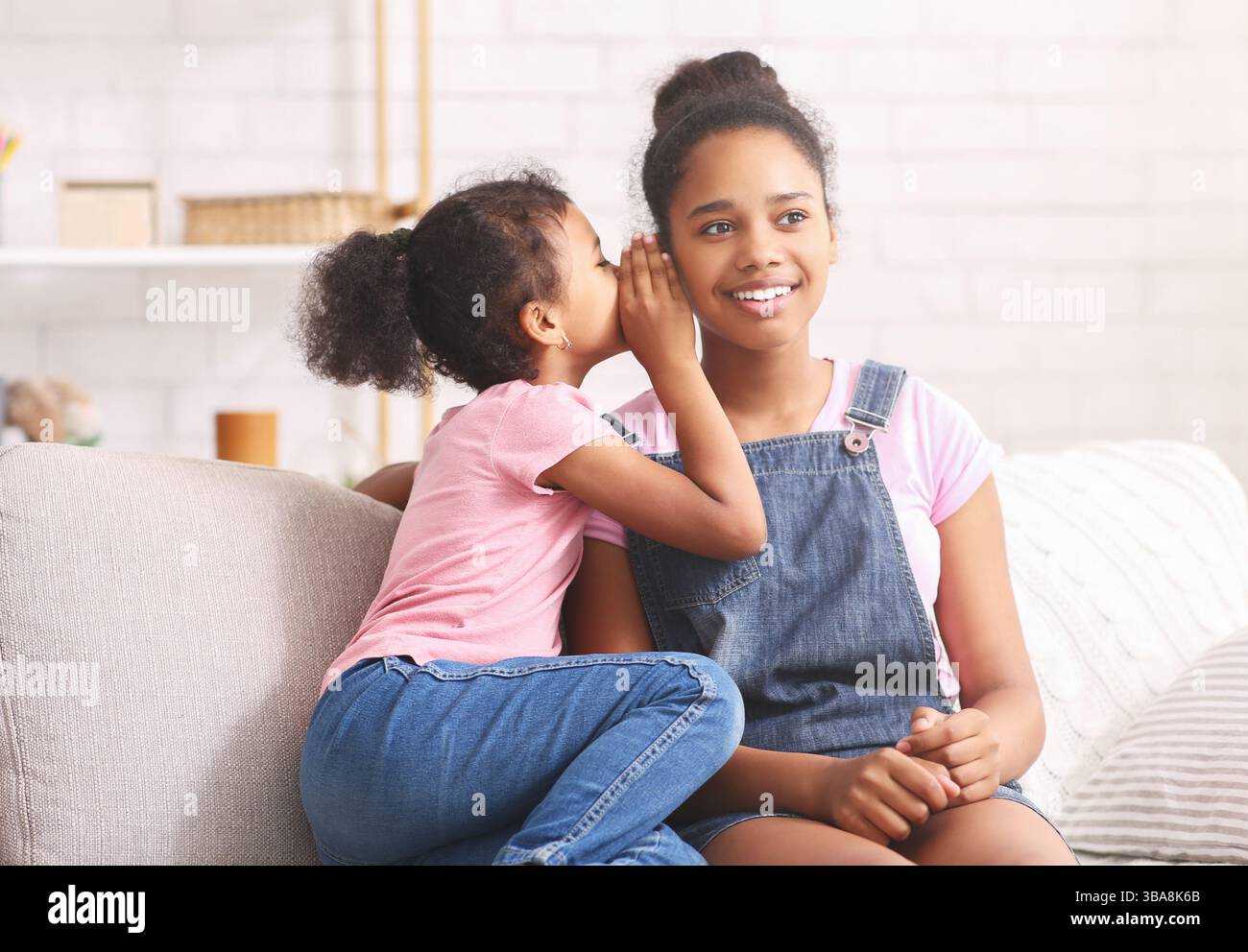 Our secret. Cute little african girl whispering something to her elder sister ear, empty space ...