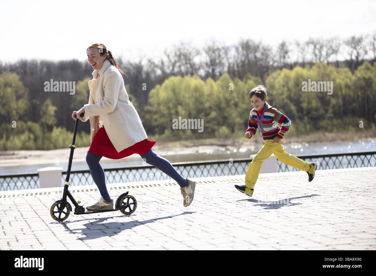 A woman rides a scooter and her son runs after her. Mother and son on a ...