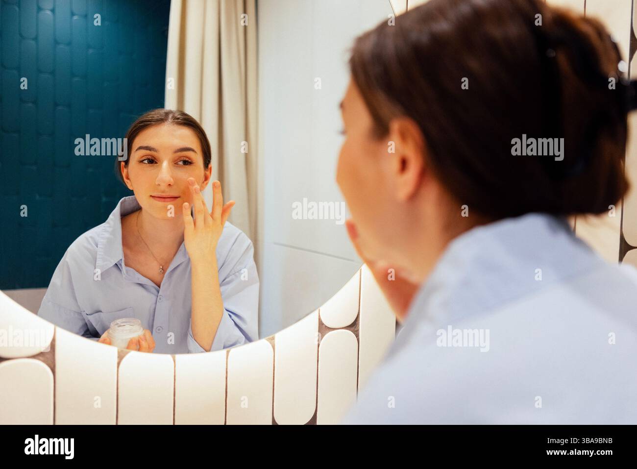 A cute girl applies a moisturizer on her face. Reflection in the ...