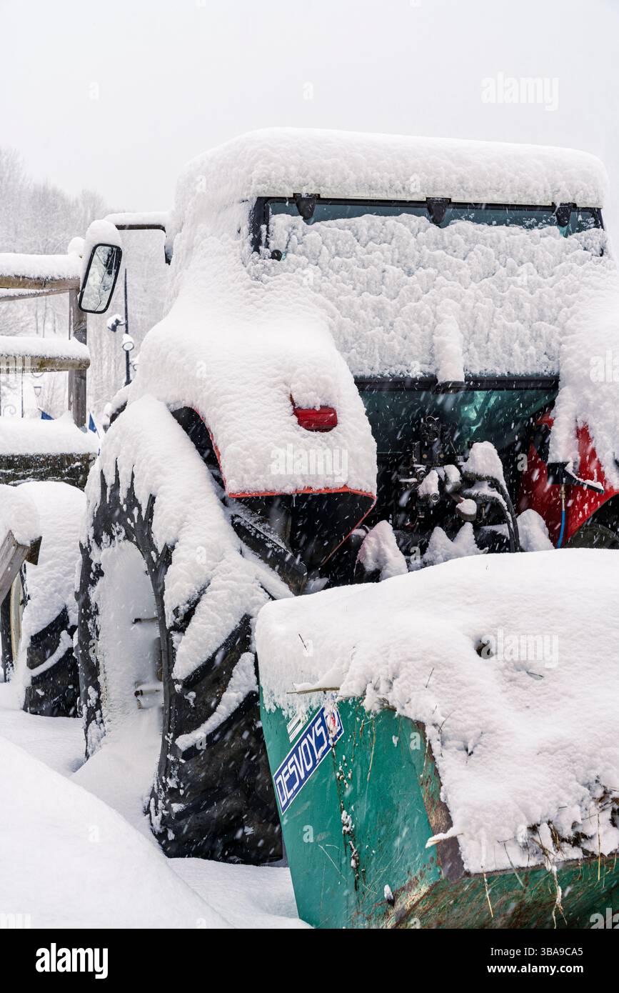 A snow-laden tractor sits idle in a village alley, its wheels and cab ...