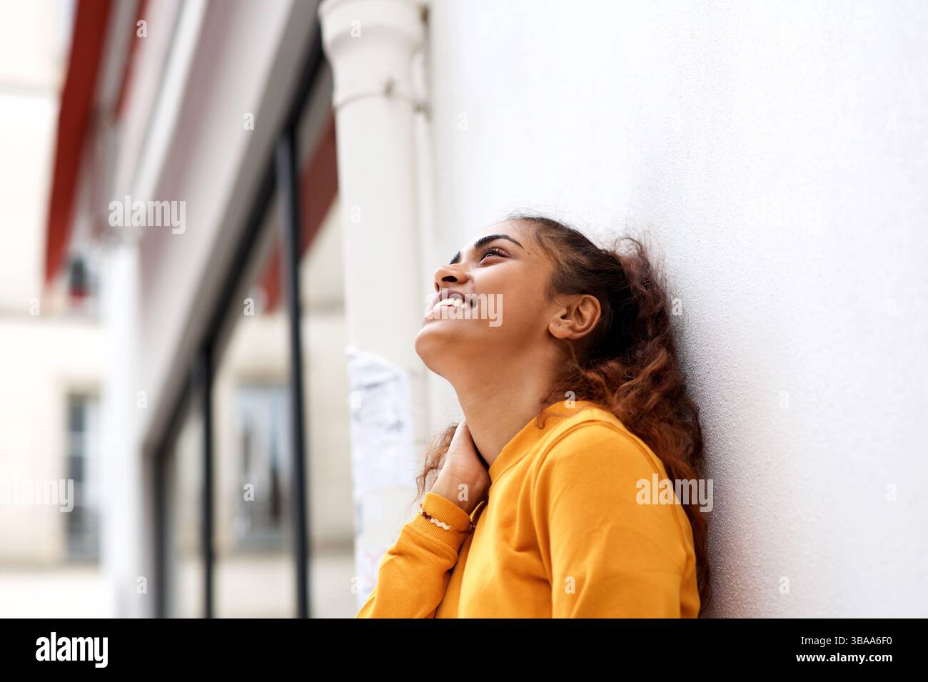 Smiling Indian woman touching neck looking up leaning against white ...