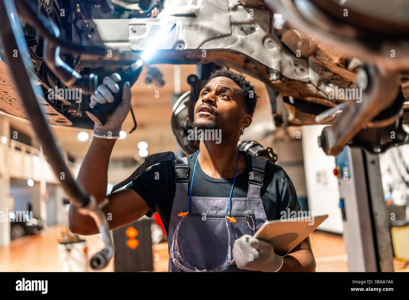 Black male mechanic inspecting car undercarriage using flashlight and holding tablet in auto ...