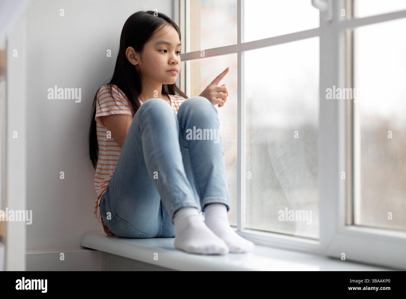Unhappy school-aged korean girl sittng on windowsill at home, drawing on glass, feeling lonely ...