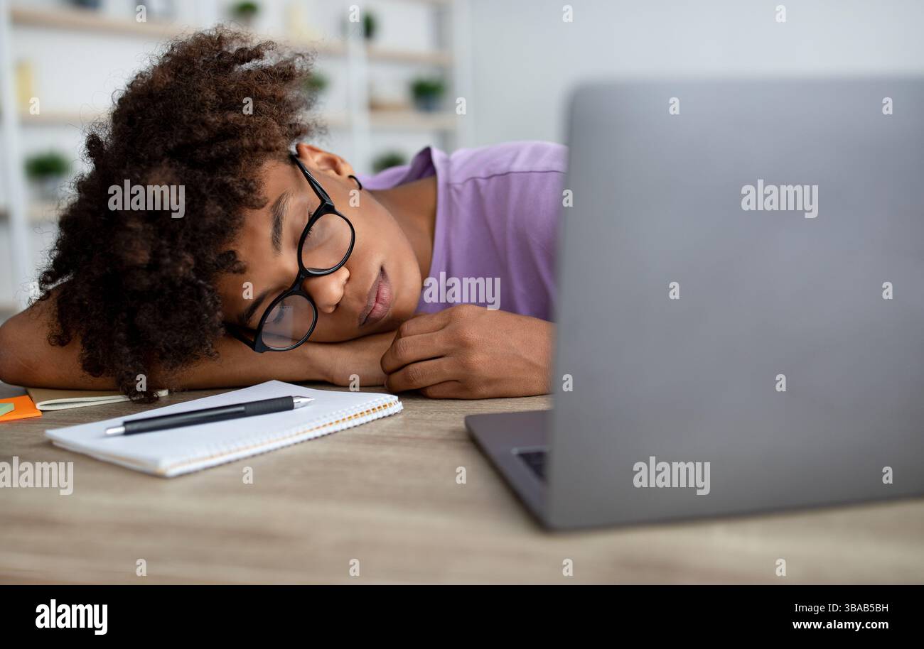 Tired black teenager sleeping on table in front of laptop computer at ...