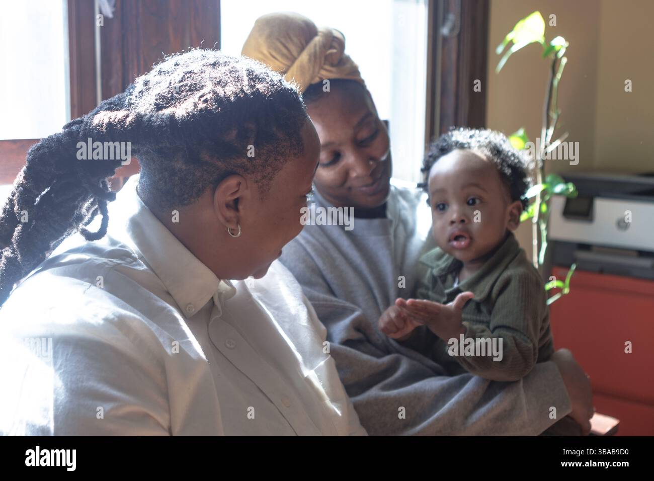 Non-binary person and Black wife playing with their baby sitting on the sofa beside the window ...