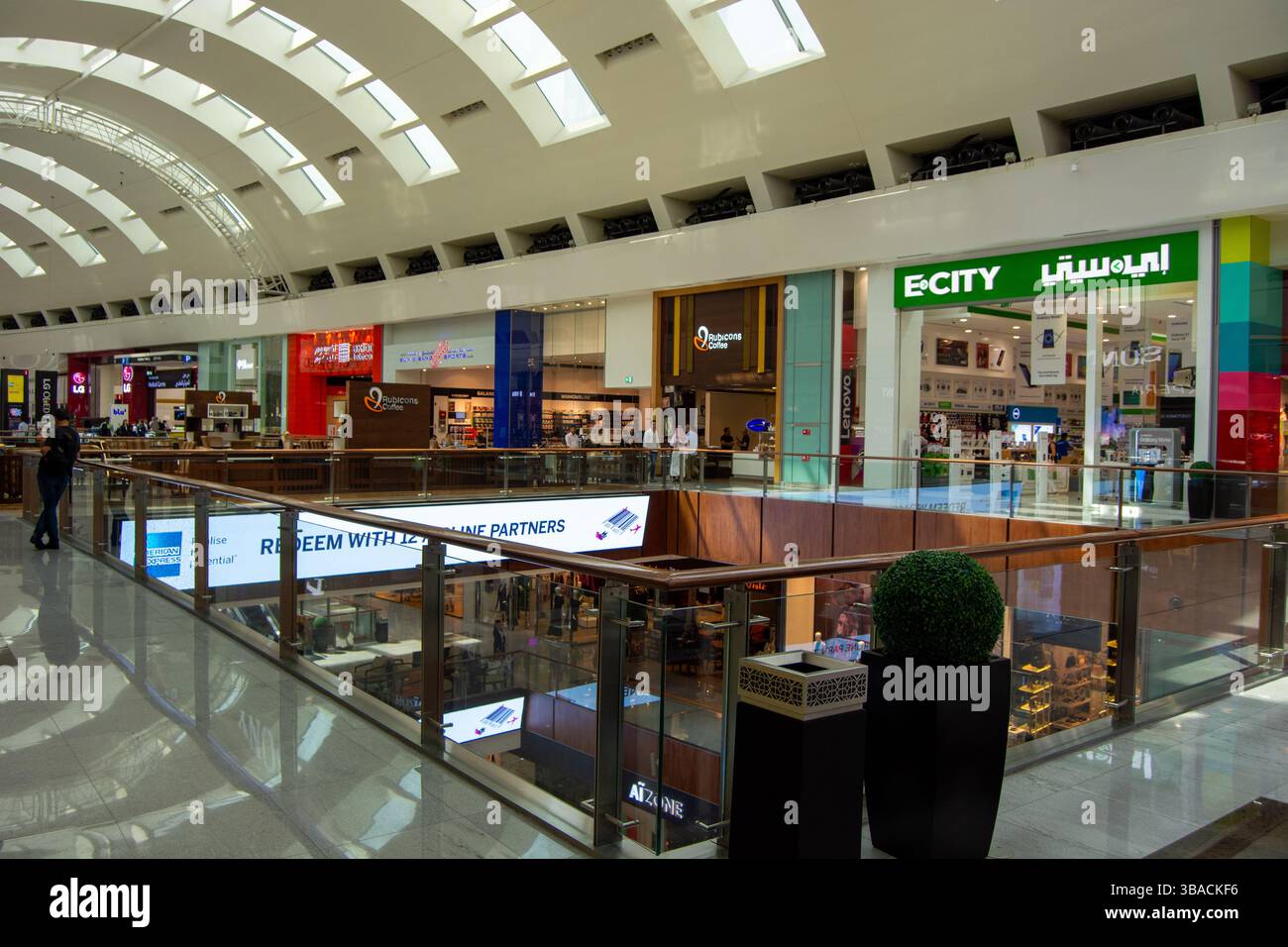 A modern shopping mall interior featuring multiple retail stores, glass railings, and bright ...