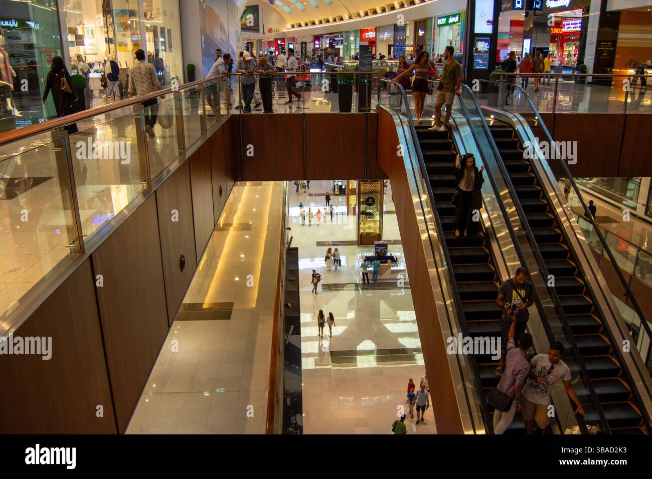 A busy shopping mall interior featuring escalators, shoppers, and various retail stores. The ...