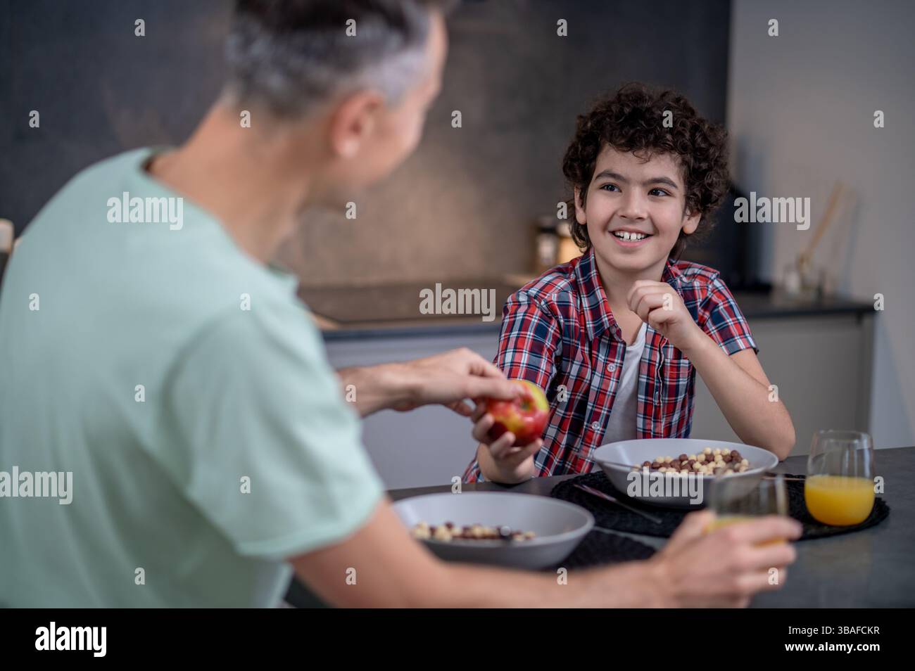 Care. Smiling boy treating father with apple sitting at table having ...