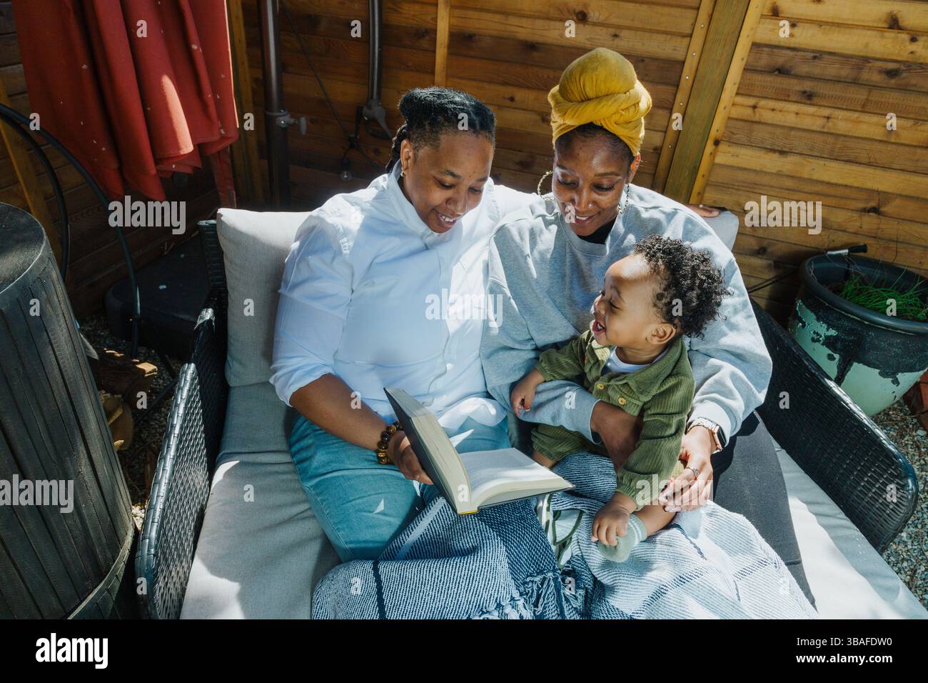 Black non-binary person with dreadlocks reading book with his wife while sitting on chair with ...