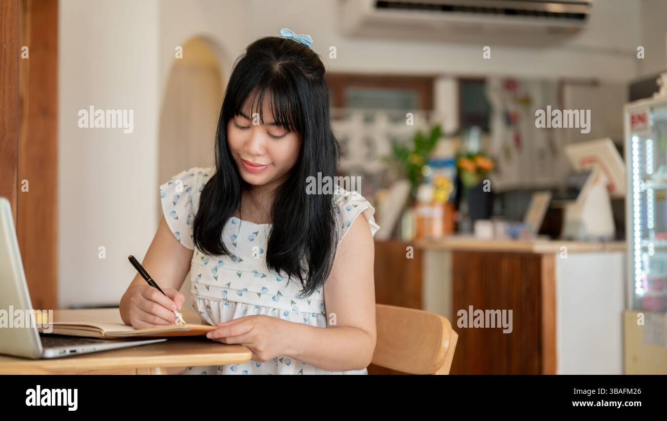 A young Asian woman focusing on writing something in a notebook or keeping diary while sitting ...
