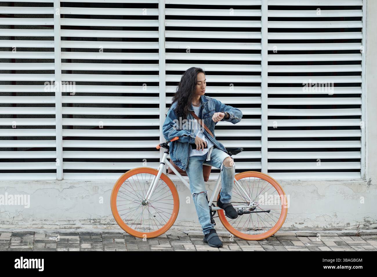 Asian man checking time on his watch while sitting on bicycle in front ...