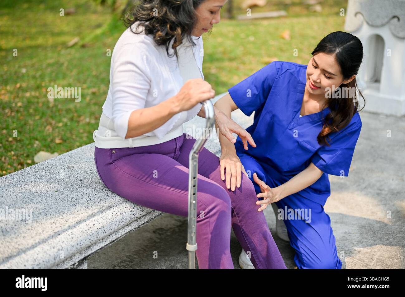 Smiling Asian female caregiver in blue uniform crouching and massaging ...