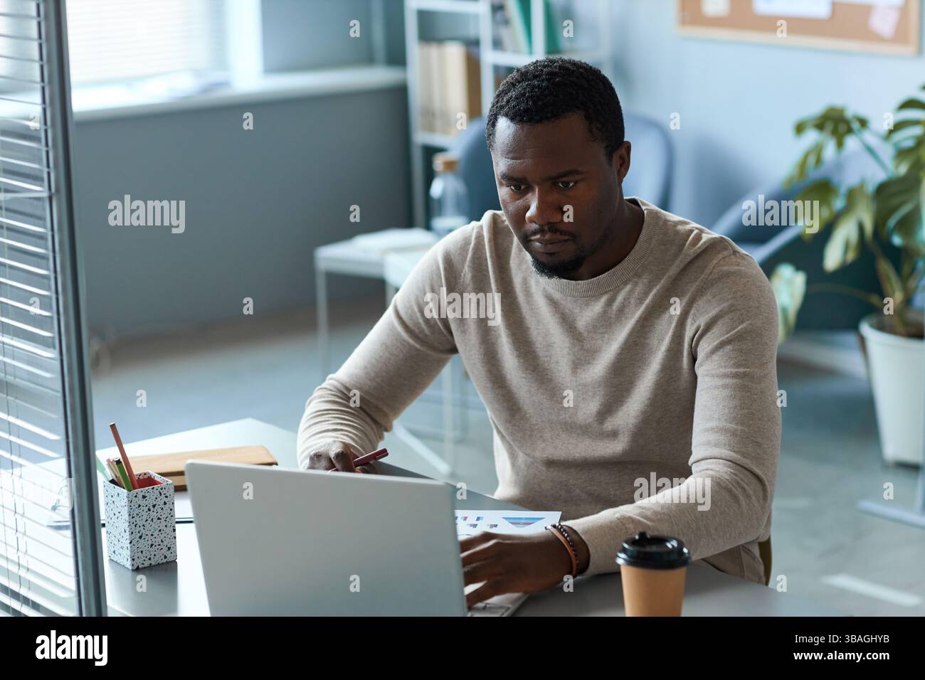 Portrait of focused black man using laptop in office behind glass partition wall, copy space ...