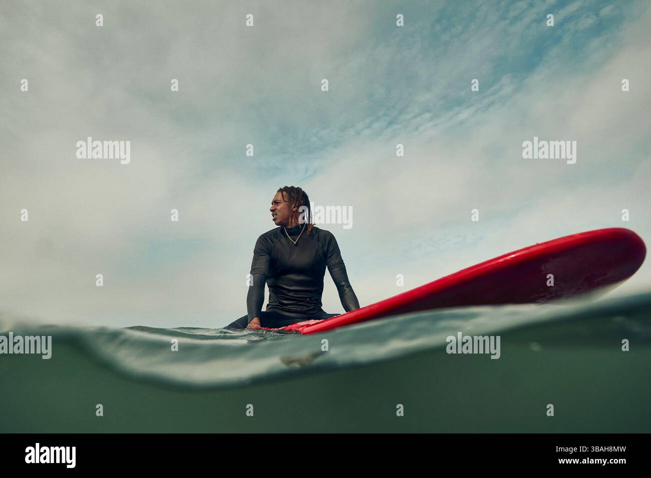 Mixed-race female surfer wearing wetsuit looking away while sitting on ...