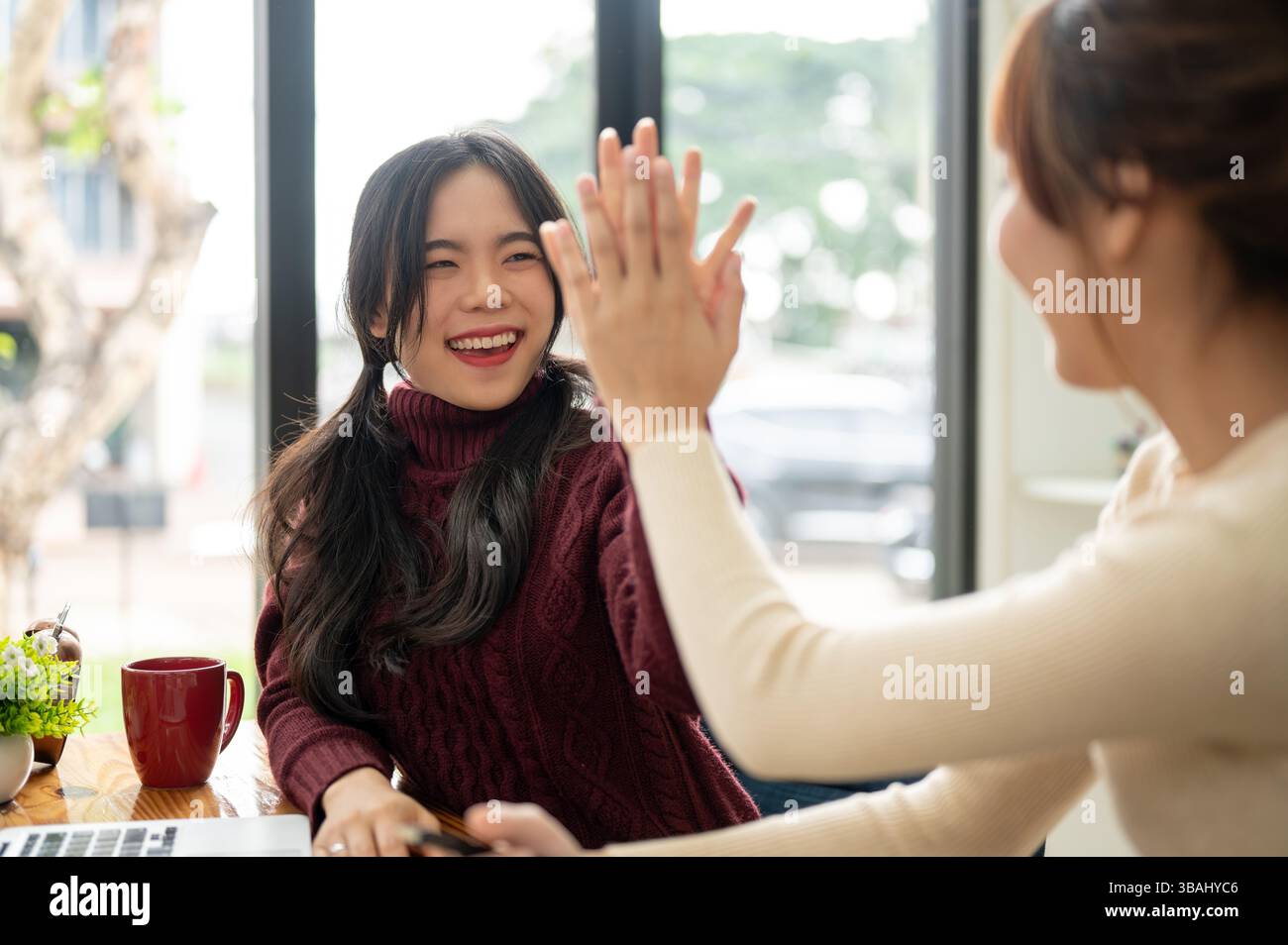 A cheerful young Asian woman is giving a high five to her friend to ...