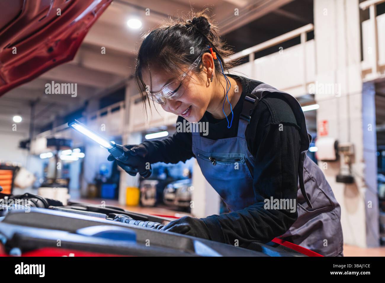 Asian woman mechanic using flashlight while inspecting car engine ...