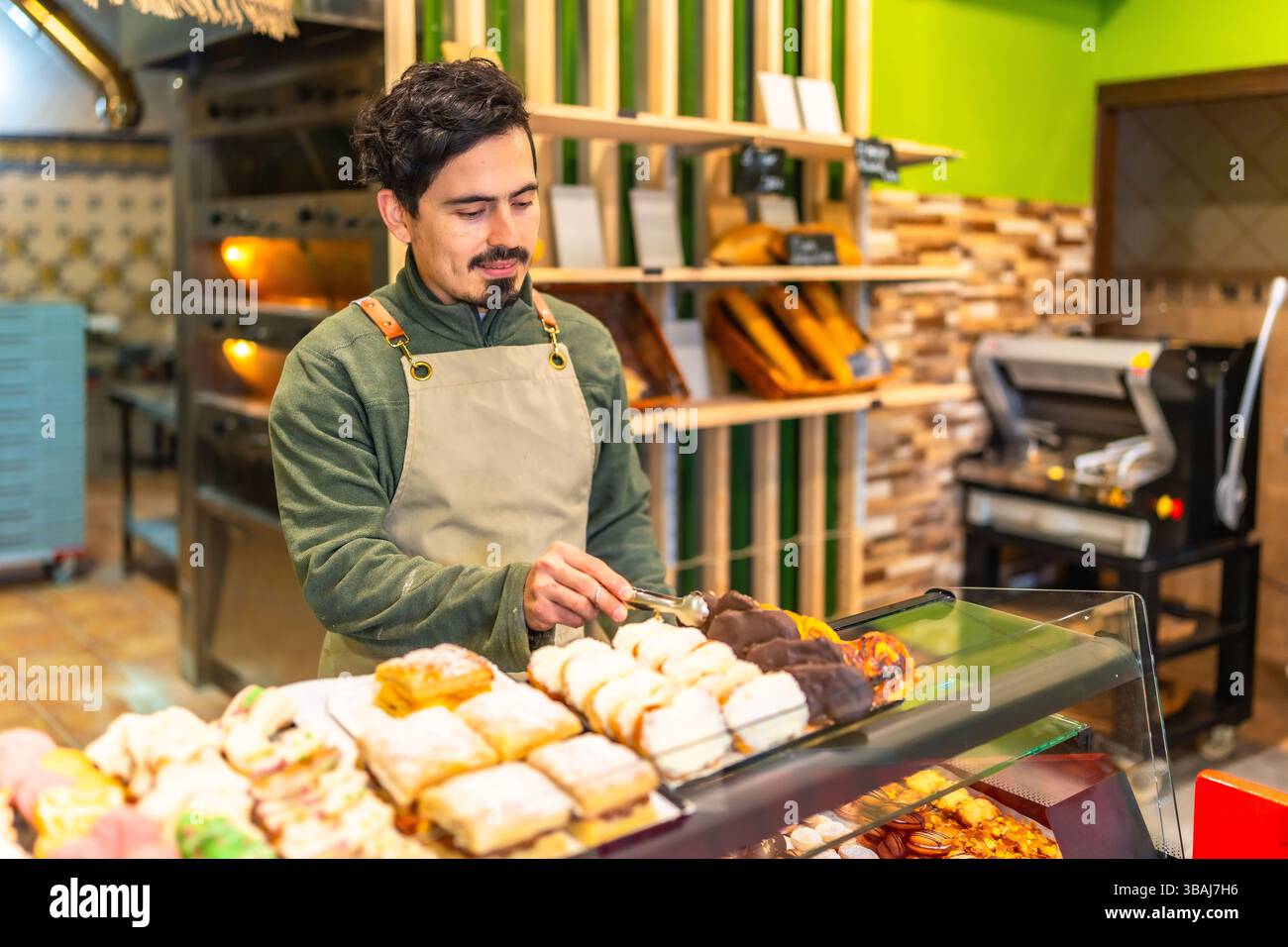 Hispanic male baker in apron arranging pastries using tong while ...