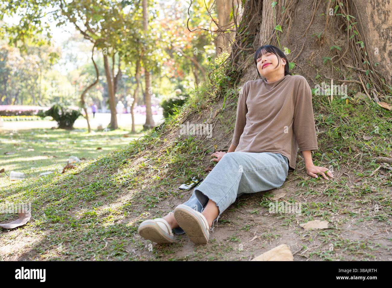 Young happy and carefree Asian woman resting under a tree in a park ...