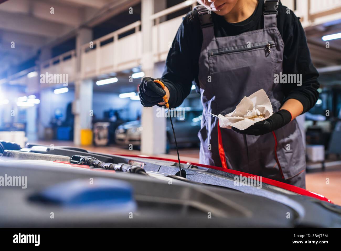 Female mechanic wearing overalls and gloves, checking the oil level of a car engine using ...