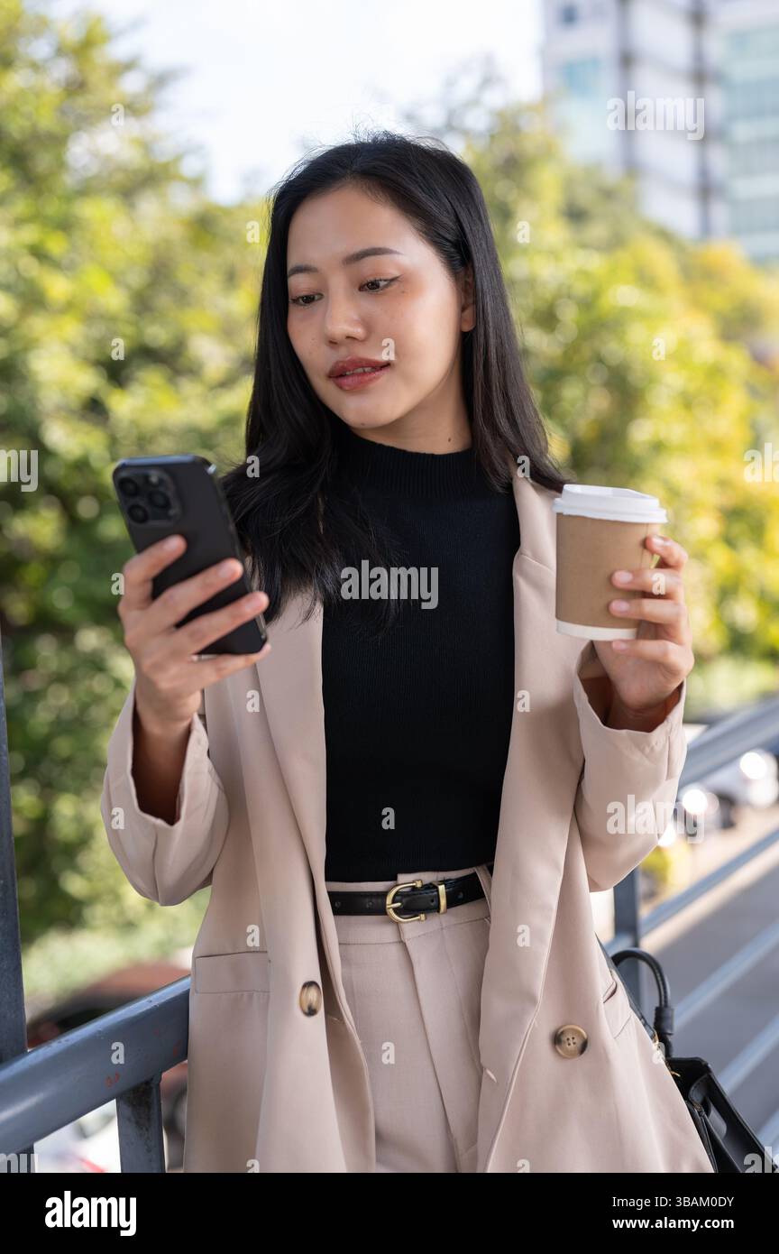 A gorgeous millennial Asian businesswoman is responding to messages on her phone on a skywalk ...