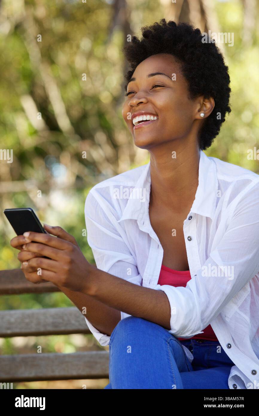 Cheerful African woman with afro hairstyle looking away holding smartphone while sitting on a ...