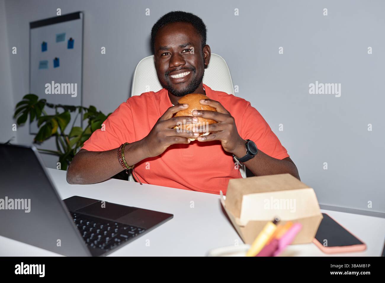 Front view portrait of smiling black man eating burger at workplace ...