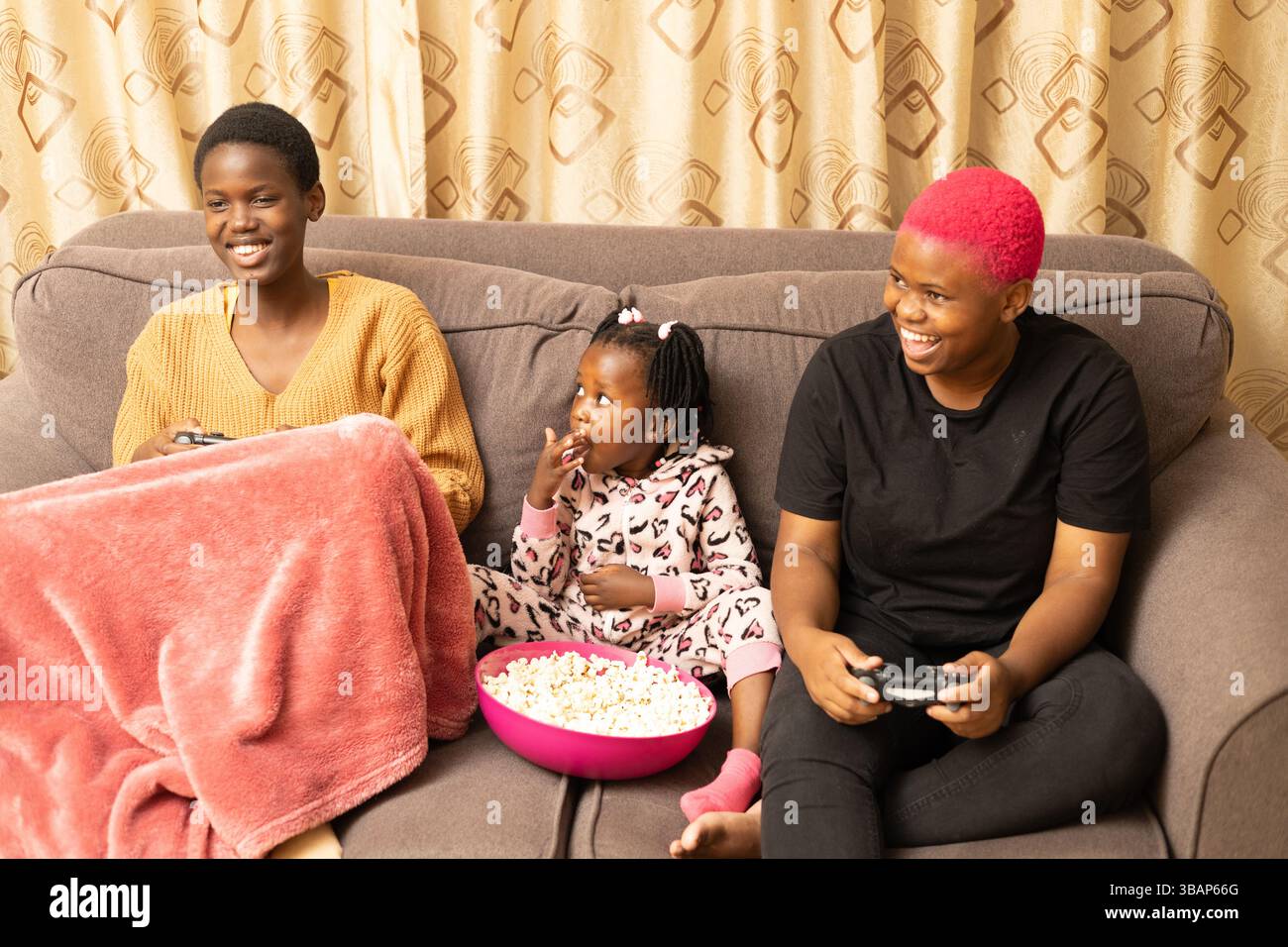 Black sisters having fun playing video games while the youngest sister is eating popcorn sitting ...