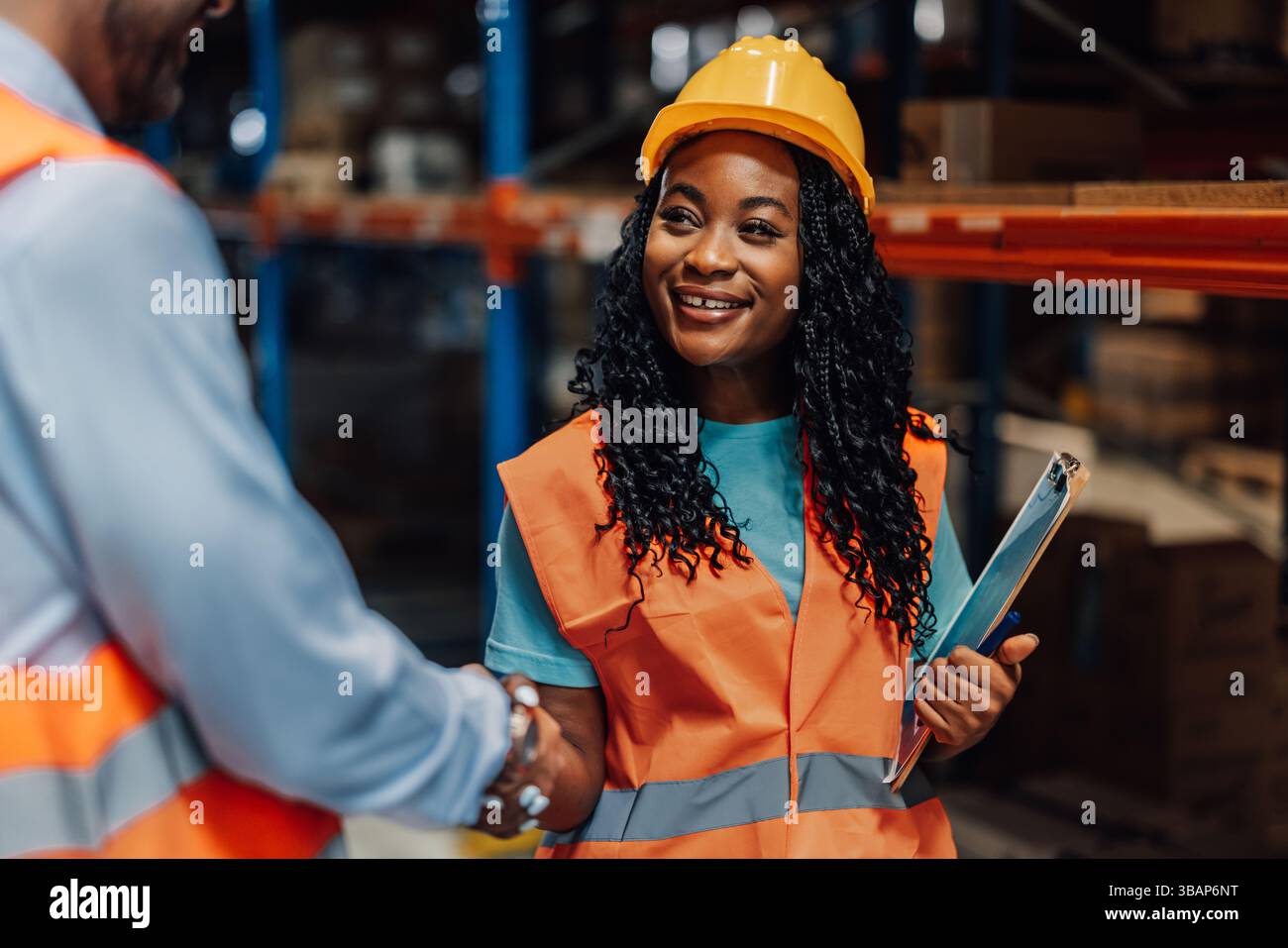 Female worker in a warehouse shakes hands with a manager, both wearing safety gear and smiling ...