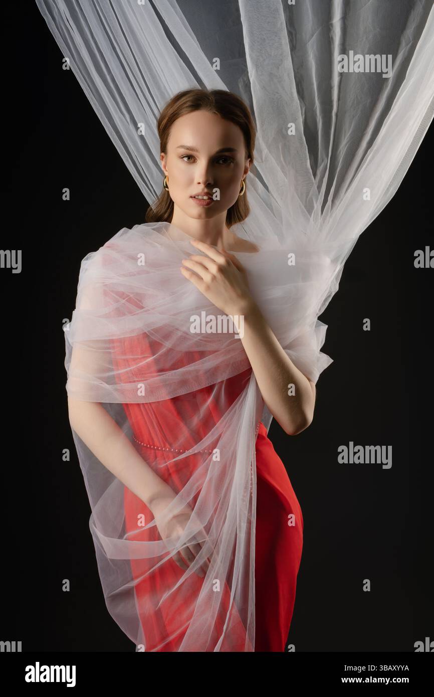 Woman poses elegantly in a studio wearing a red dress and sheer fabric ...